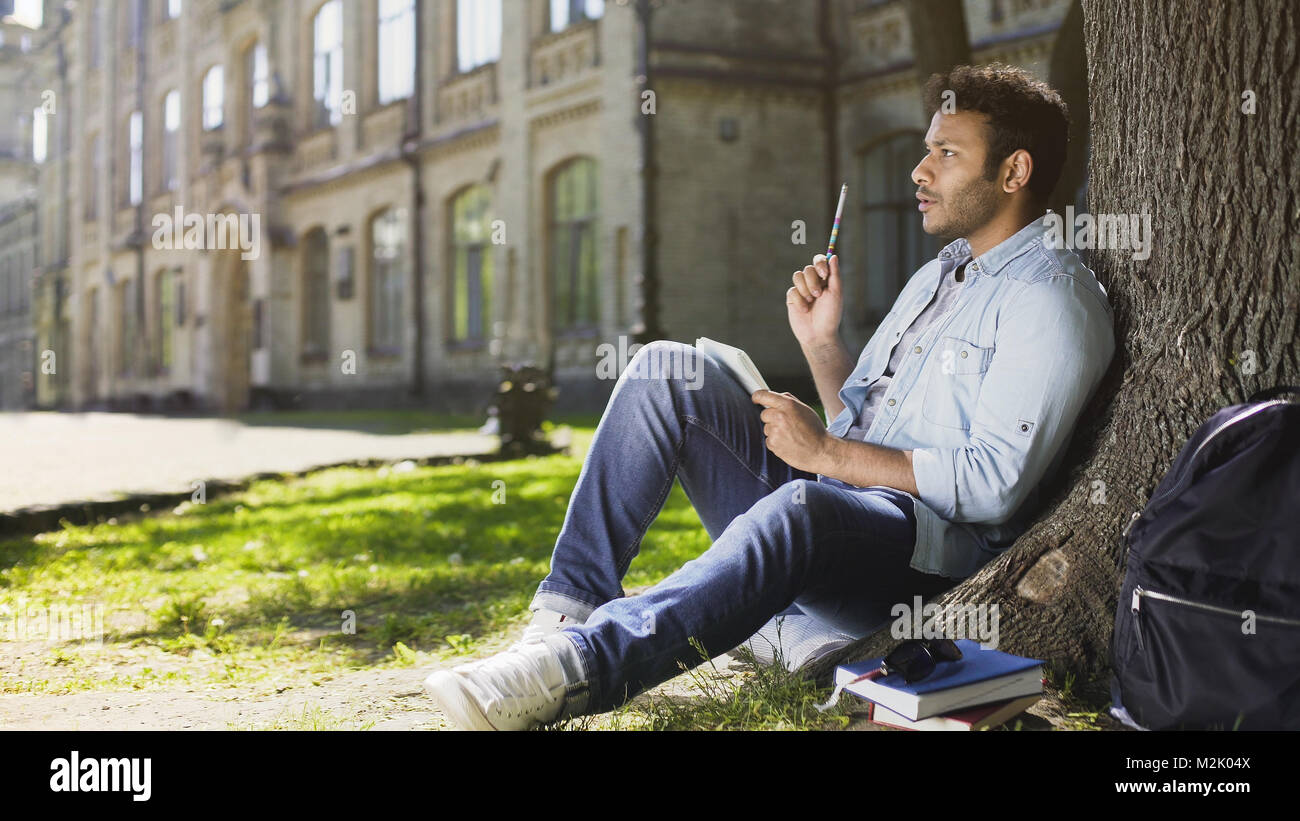 Multinational guy sitting under tree in park writing creative idea in ...