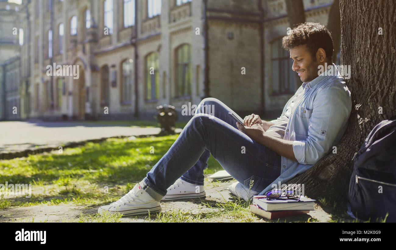 Multiracial young man sitting under tree, scrolling mobile phone screen ...
