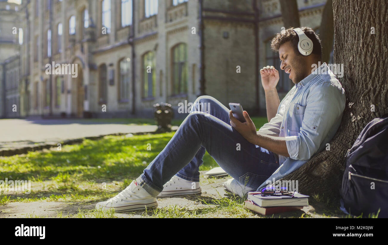 Guy in headphones sitting on grass and leaning against tree, singing to ...