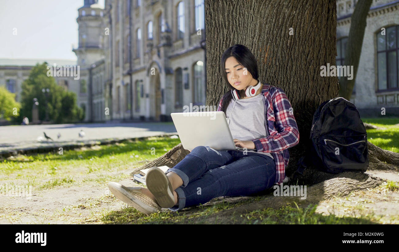 College student sitting under tree in campus, using laptop, writing ...