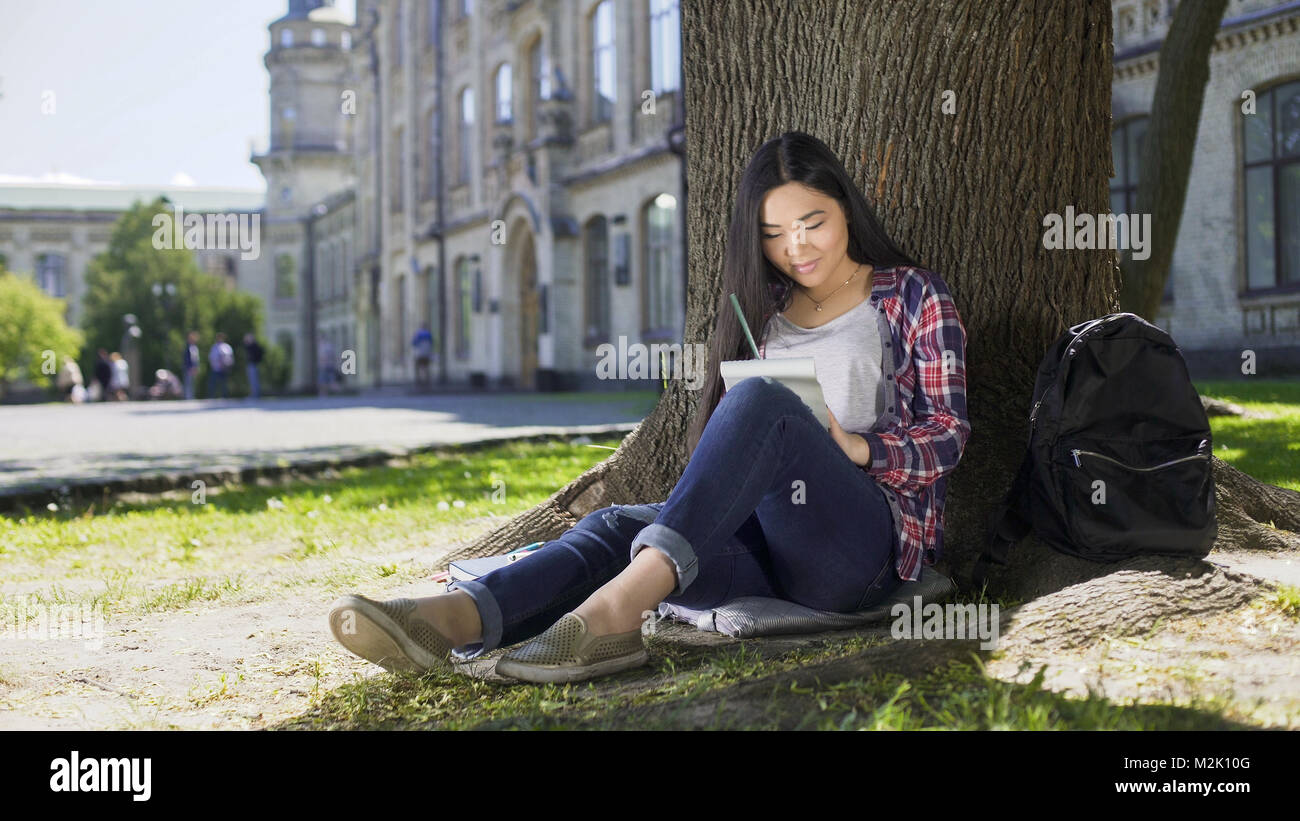 Multinational young woman taking notes in notebook sitting under tree ...