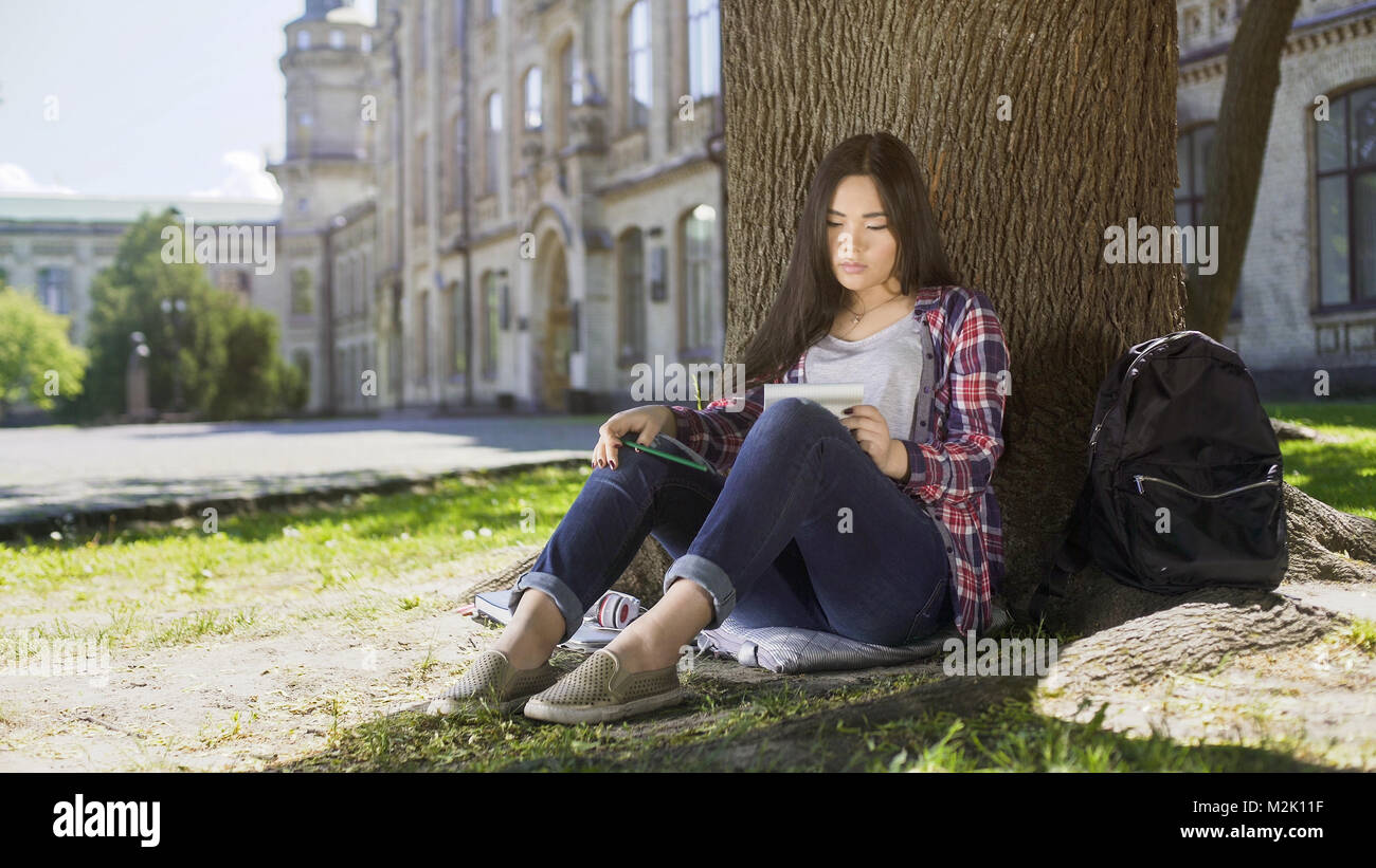 International student sitting under tree, holding notebook, looking at ...