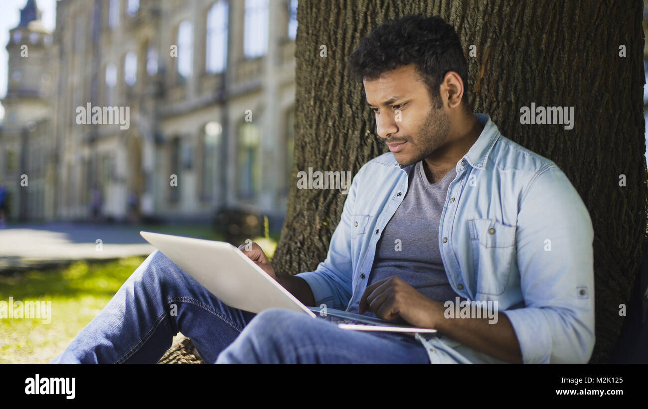 Multiracial college student sitting under tree with laptop, checking ...