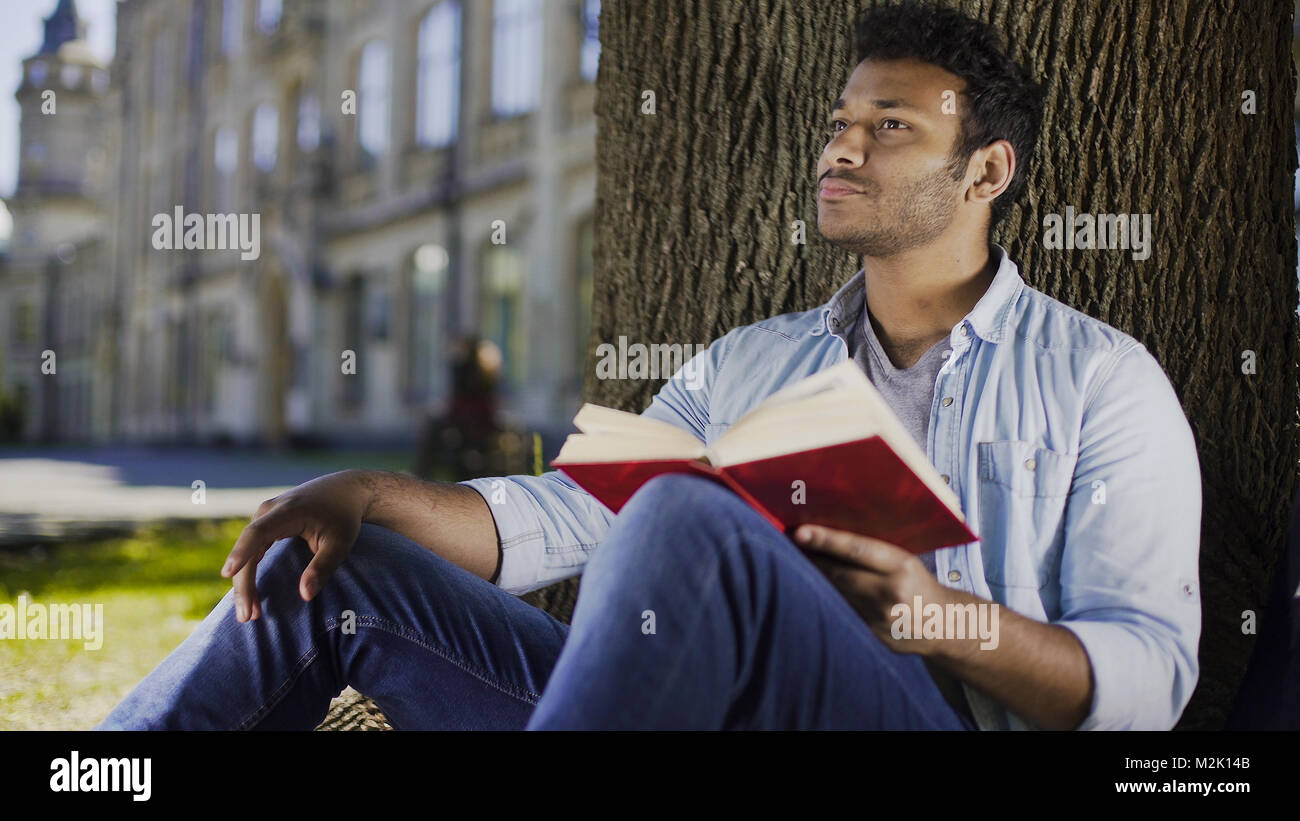 Young Man Sitting Under Tree Reading Book And Thinking Fiction