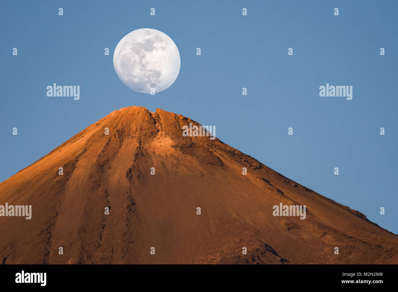 The full moon rising above the peak of Mount Teide against a blue sky ...