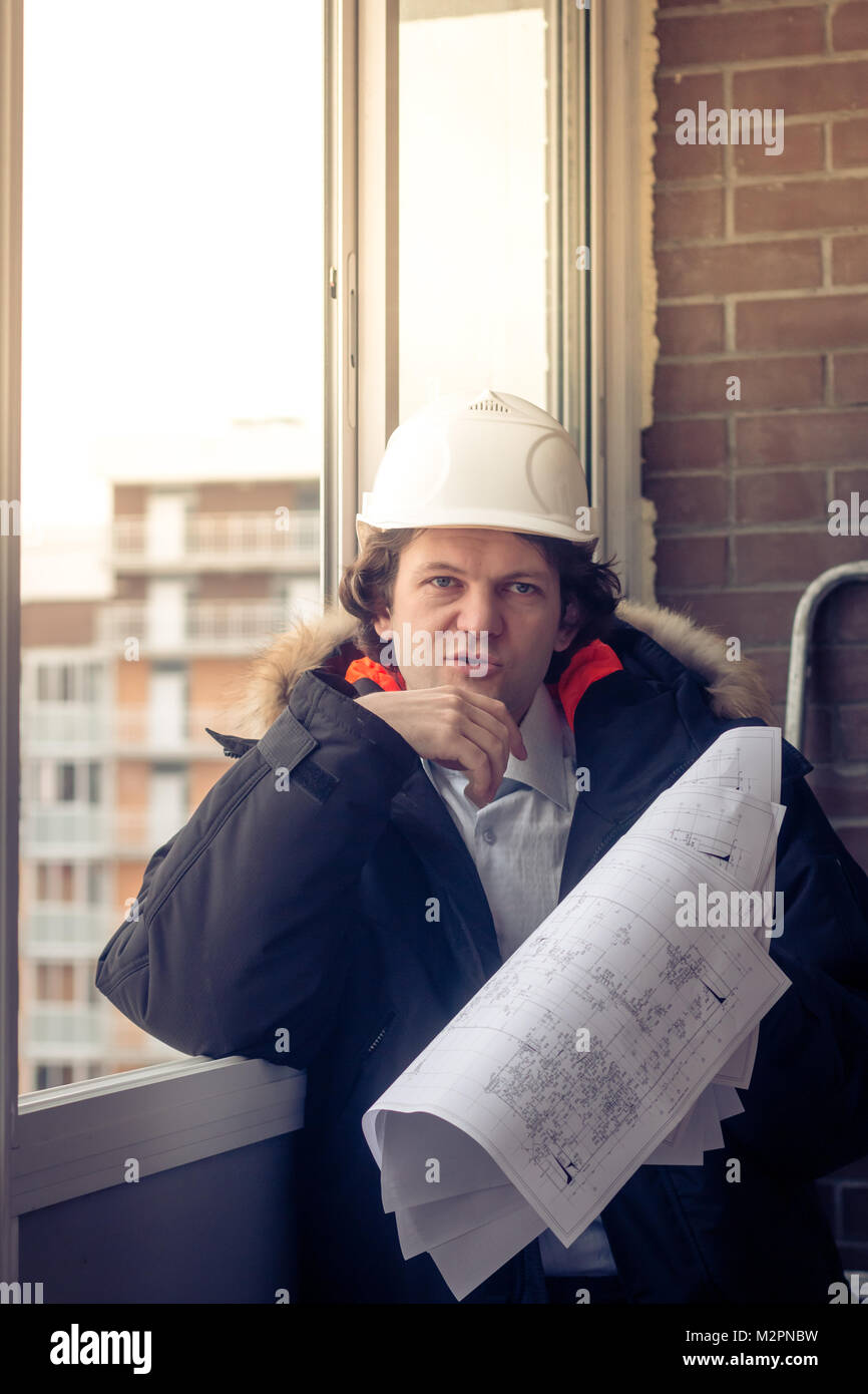 Pensive young man builder in hard hat standng and thinking. Soft focus ...