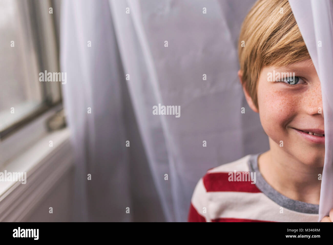 Portrait of a smiling boy with freckles standing by a window hiding ...