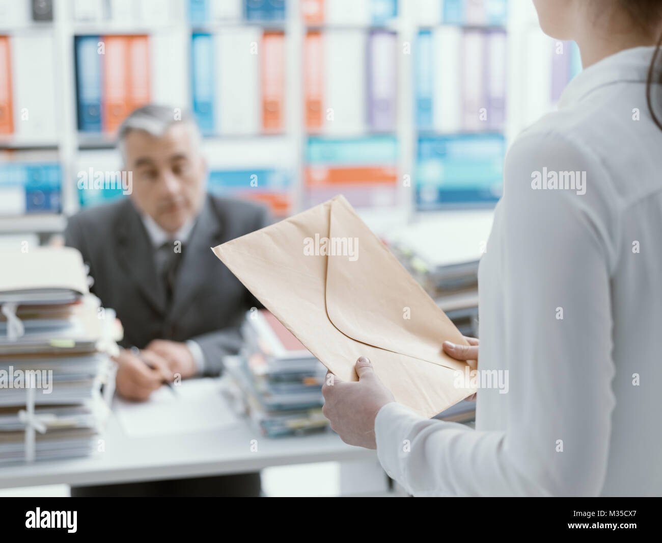 Assistant bringing mail to her boss, he is working at desk overloaded ...