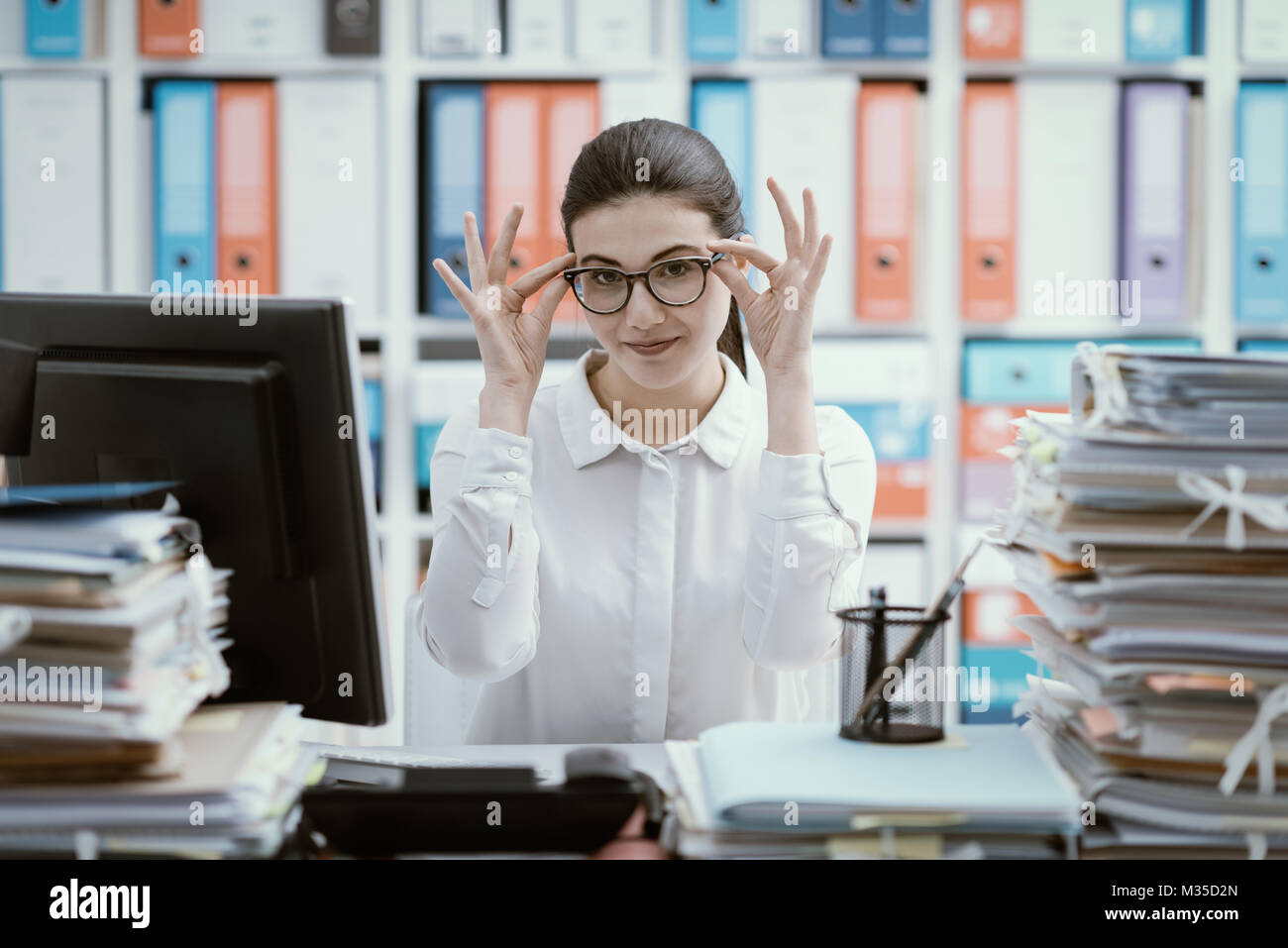 Confident secretary sitting at office desk and adjusting her glasses ...