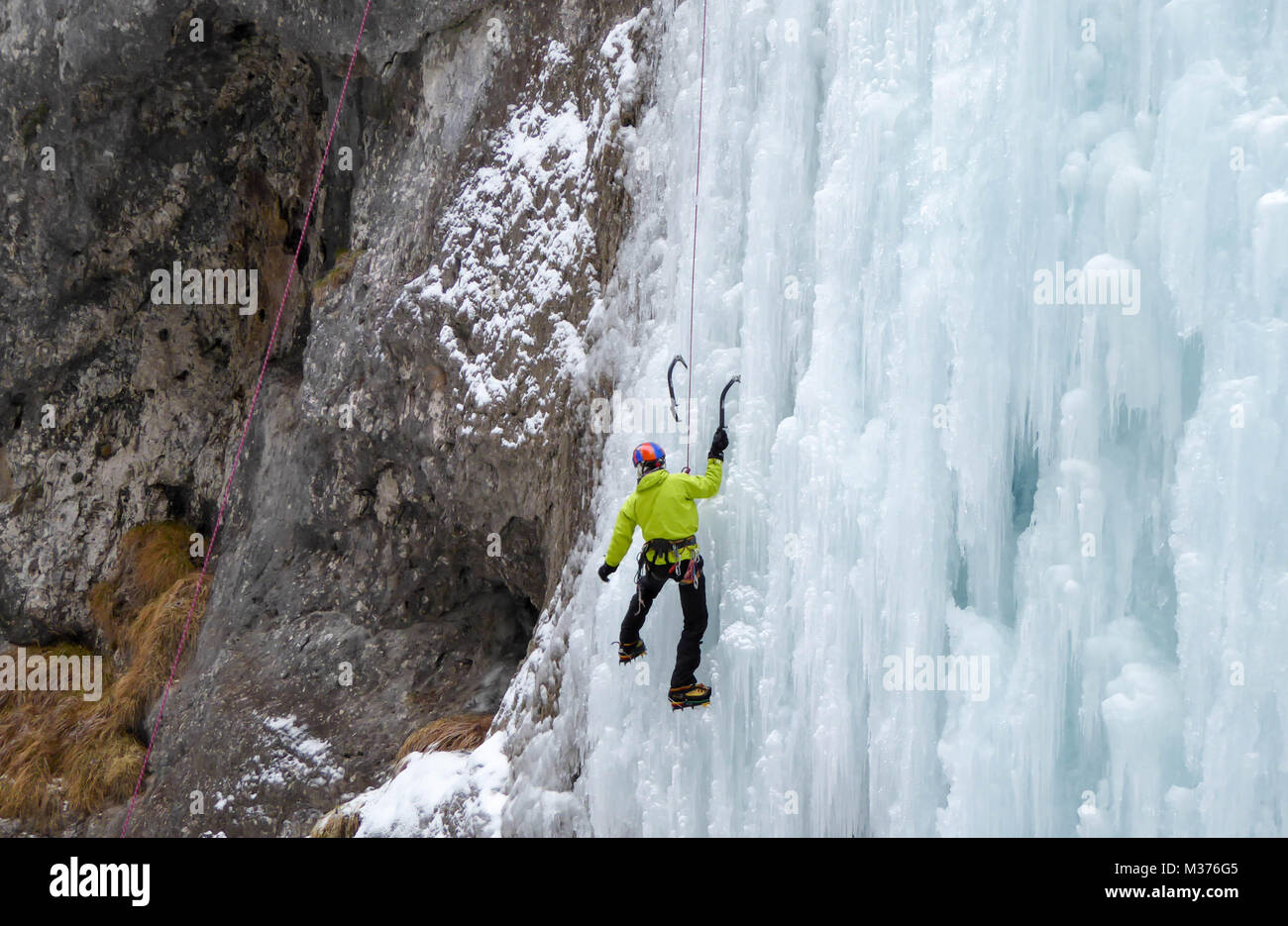 male ice climber in a yellow jacket on a steep frozen waterfall ice ...