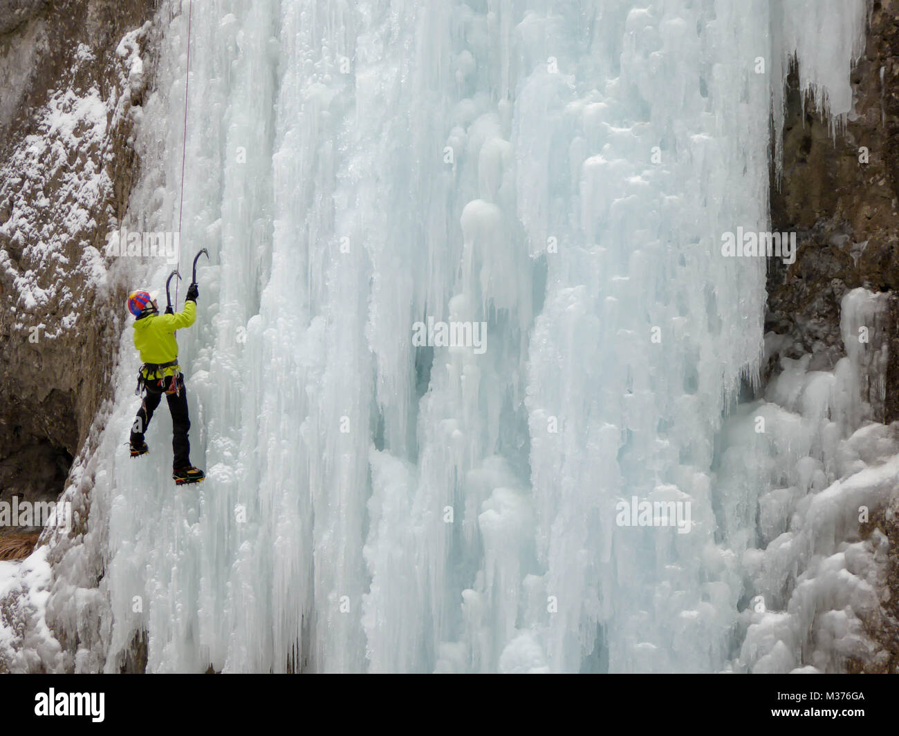 male ice climber in a yellow jacket on a steep frozen waterfall ice ...