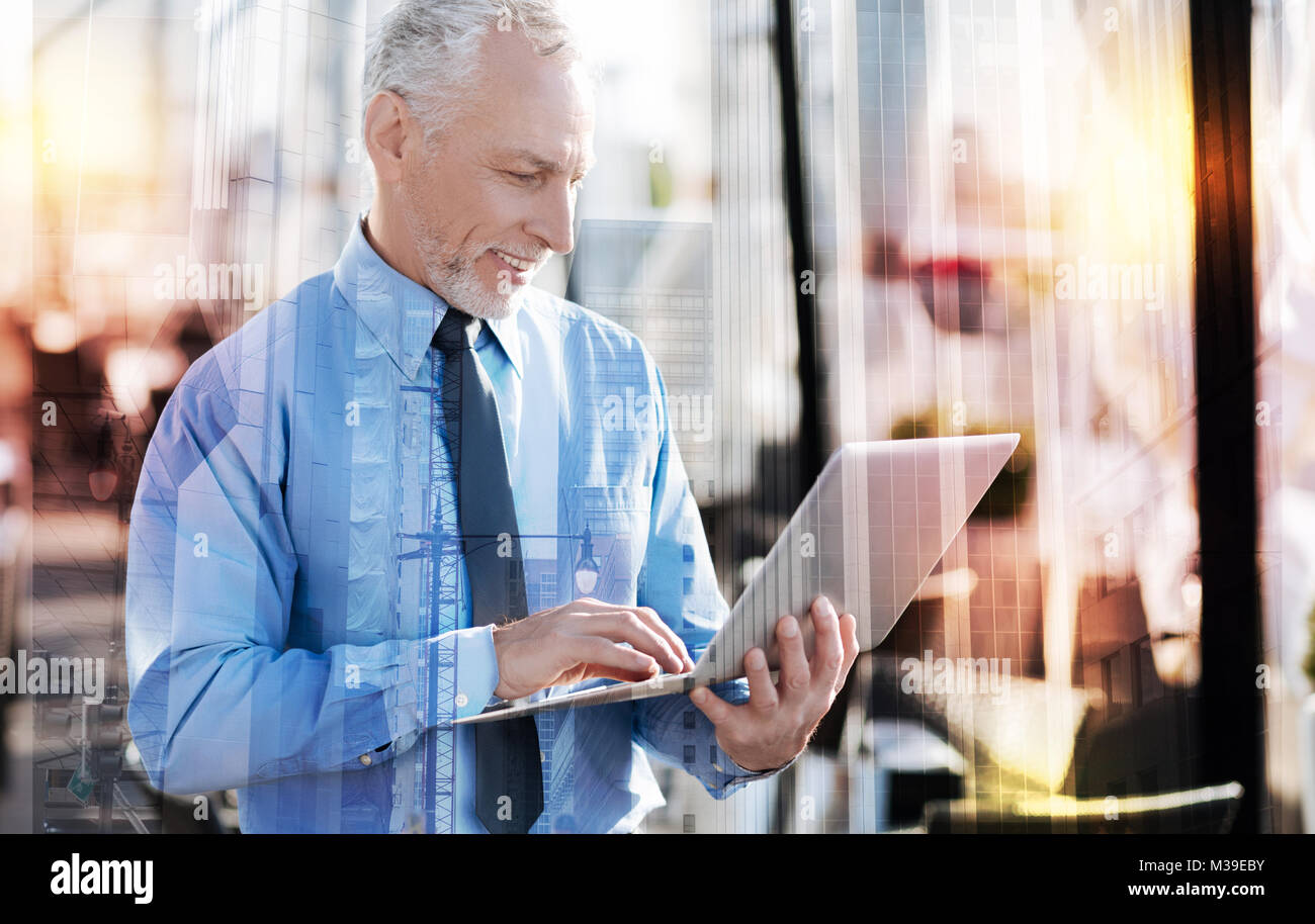 Progressive mature man smiling while standing with a modern laptop ...