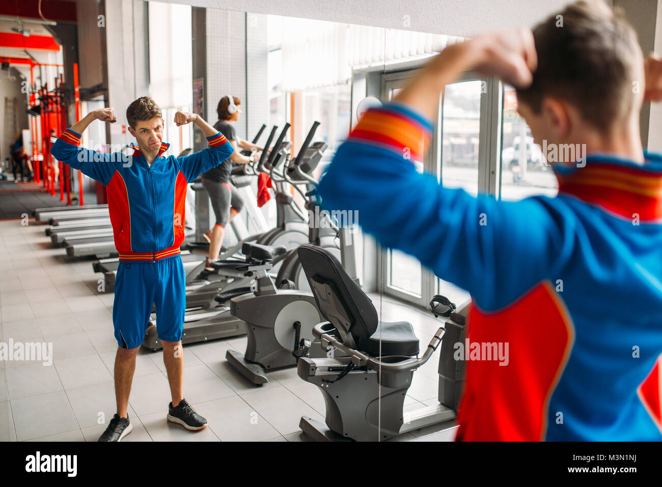 Skinny man in sportswear watches on his muscles in mirror at the gym ...