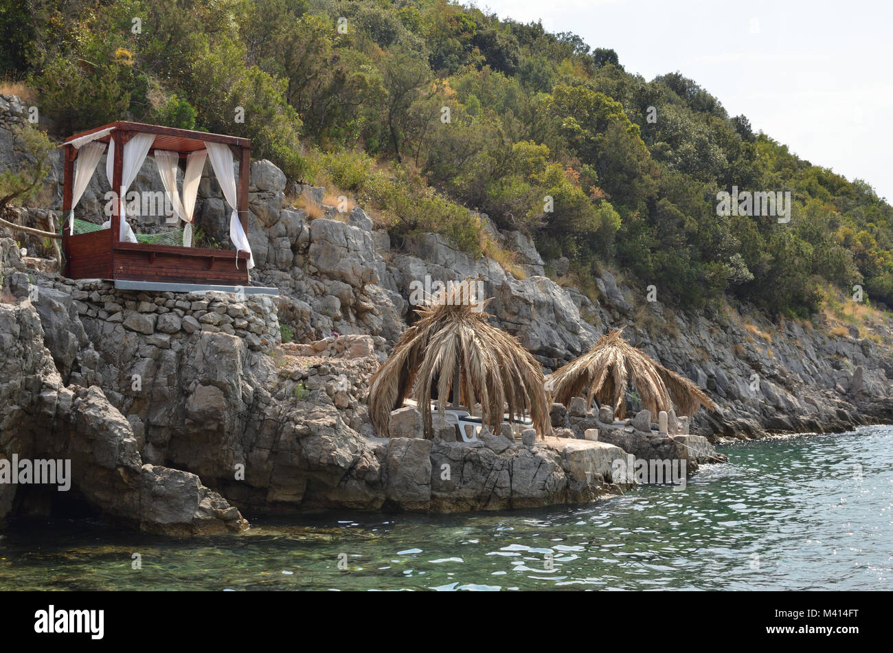 Straw parasols and cabana bed on beach rocks with green sea water in ...