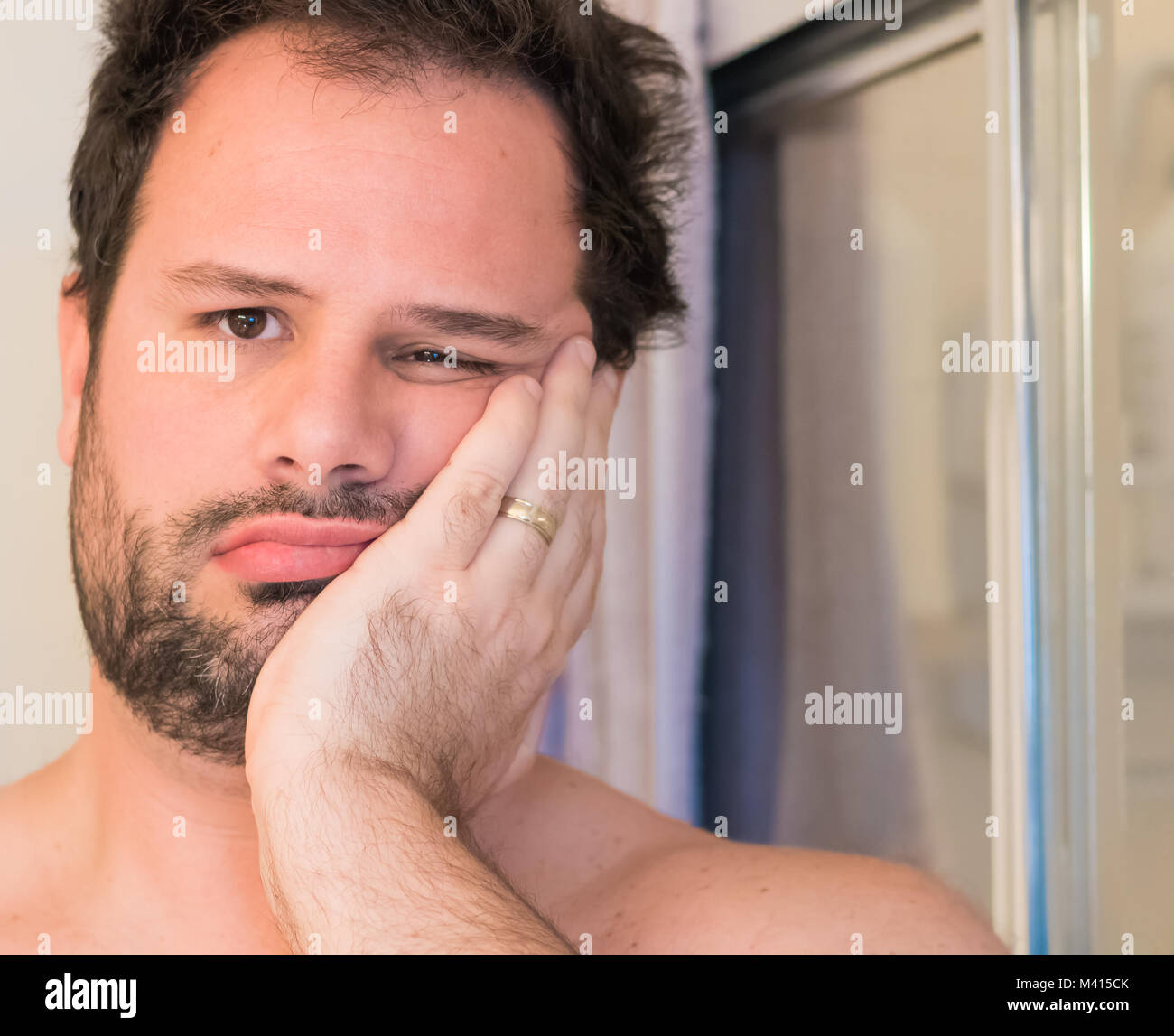 A young adult Caucasian male has his hand on his cheek as he hesitates ...