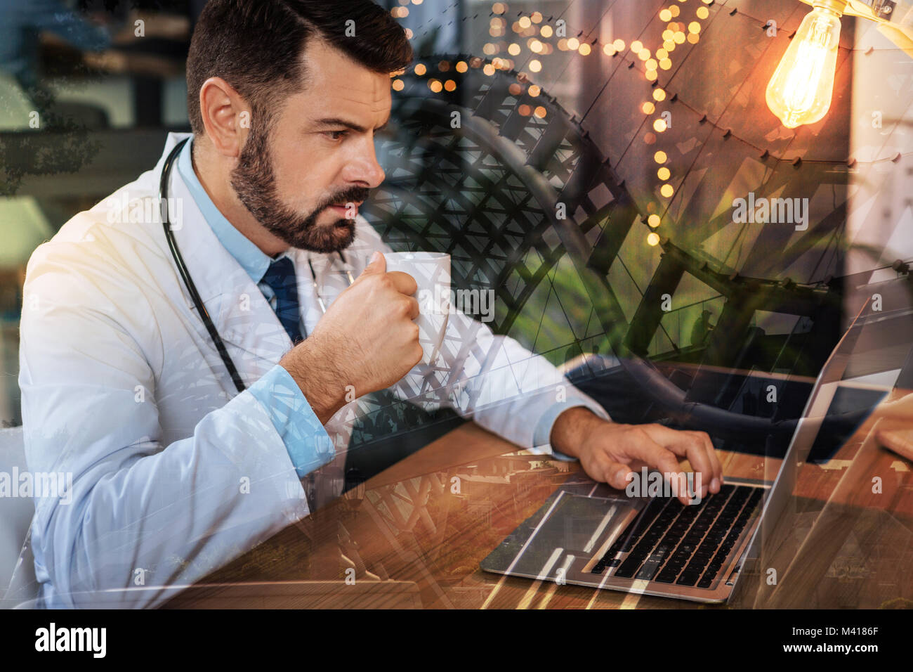 Calm doctor drinking coffee while looking at the screen of a laptop ...
