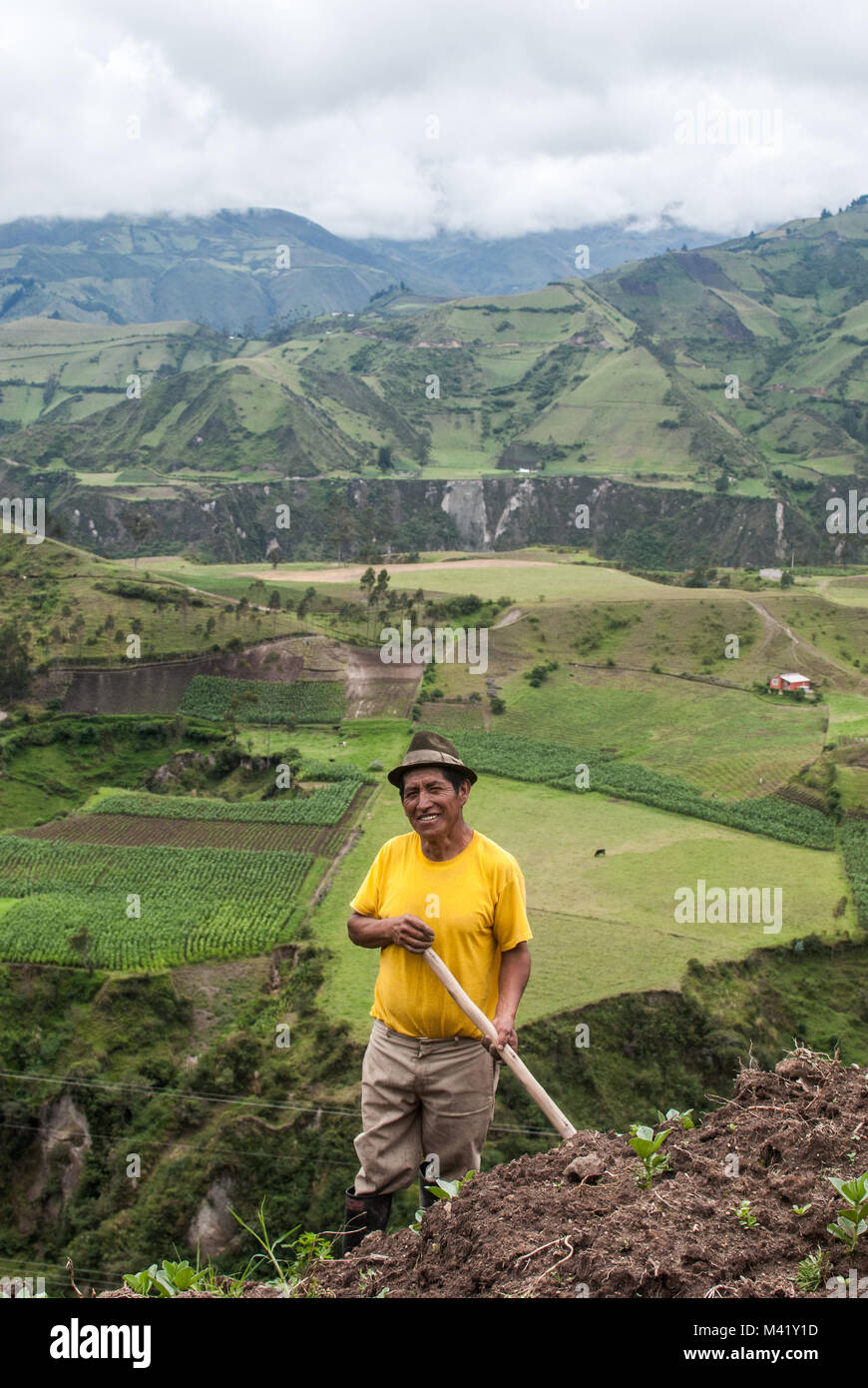 A peasant farmer smiling and holding a tool in front of a mountain ...