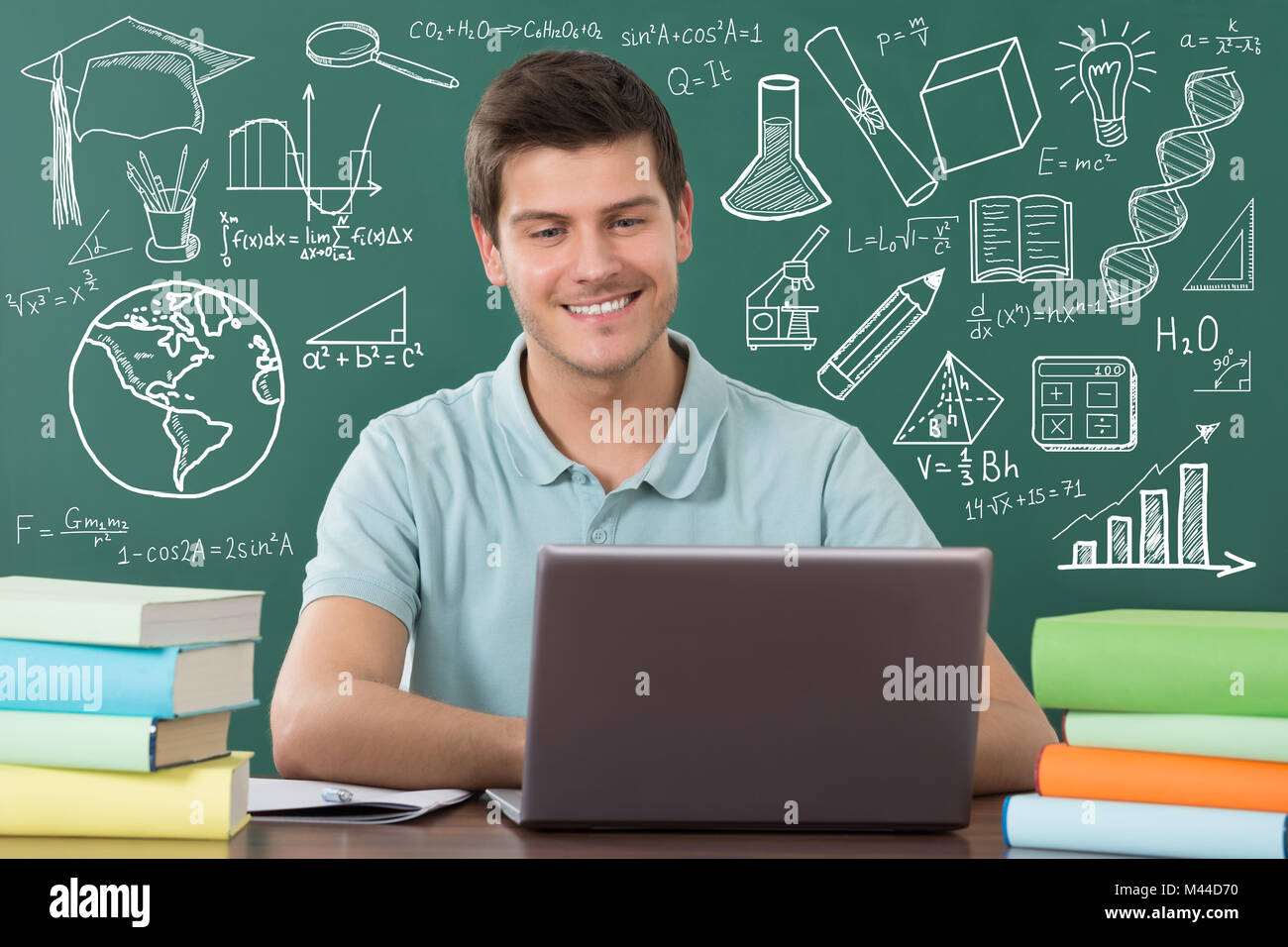 Smiling Male Student Using Laptop Against The Green Chalkboard In ...