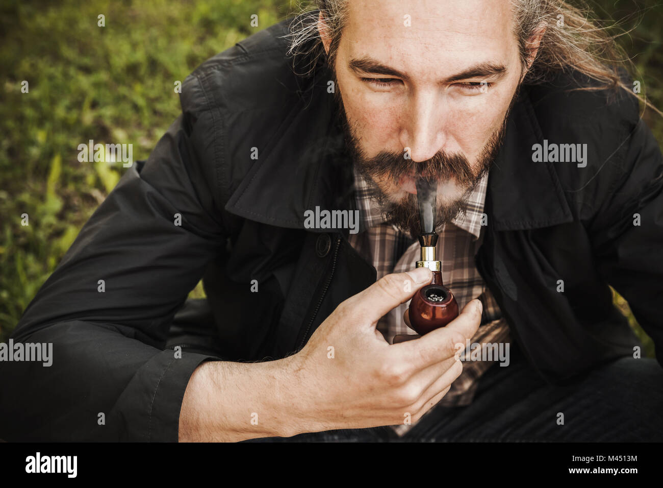 Young serious bearded man smoking pipe in summer park, close up ...