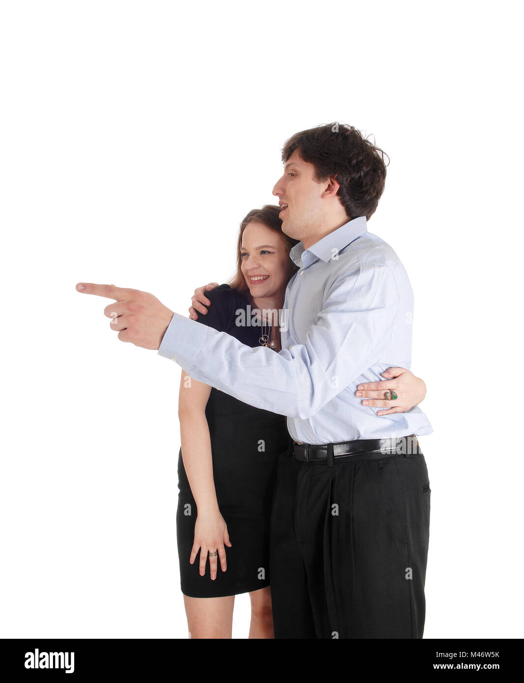 A young couple standing, she is hugging him, he is pointing away and ...