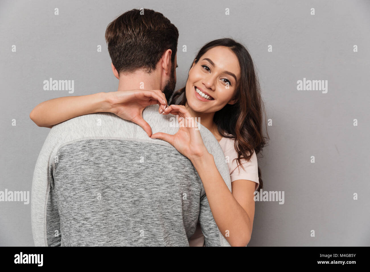 Portrait of a happy young woman hugging her boyfriend and heart gesture with fingers over gray ...