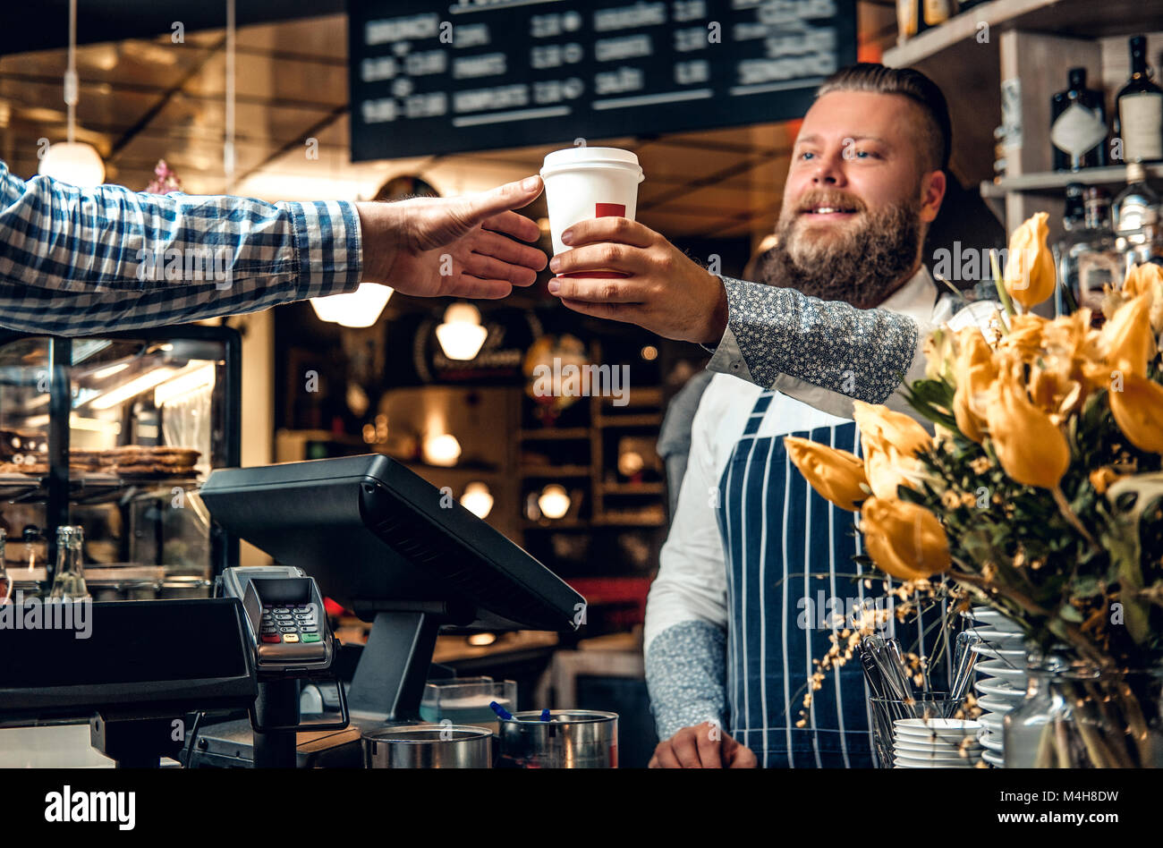 Positive bearded barista male selling coffee to a consumer in a coffee ...