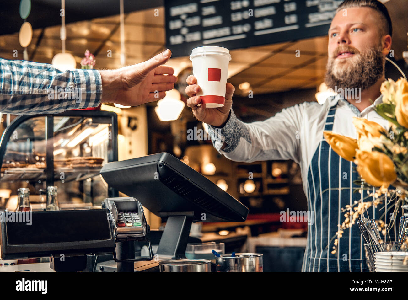 Positive bearded barista male selling coffee to a consumer in a coffee ...