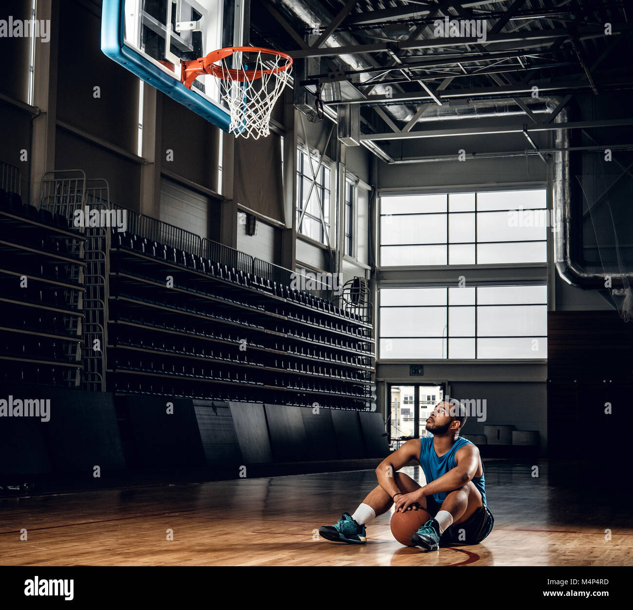 Portrait of Black basketball player sits on a floor under a hoop in a ...