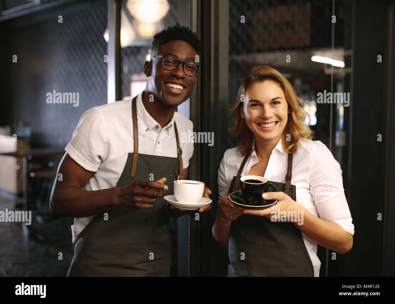 Male and female barista standing at the entrance of coffee shop holding ...