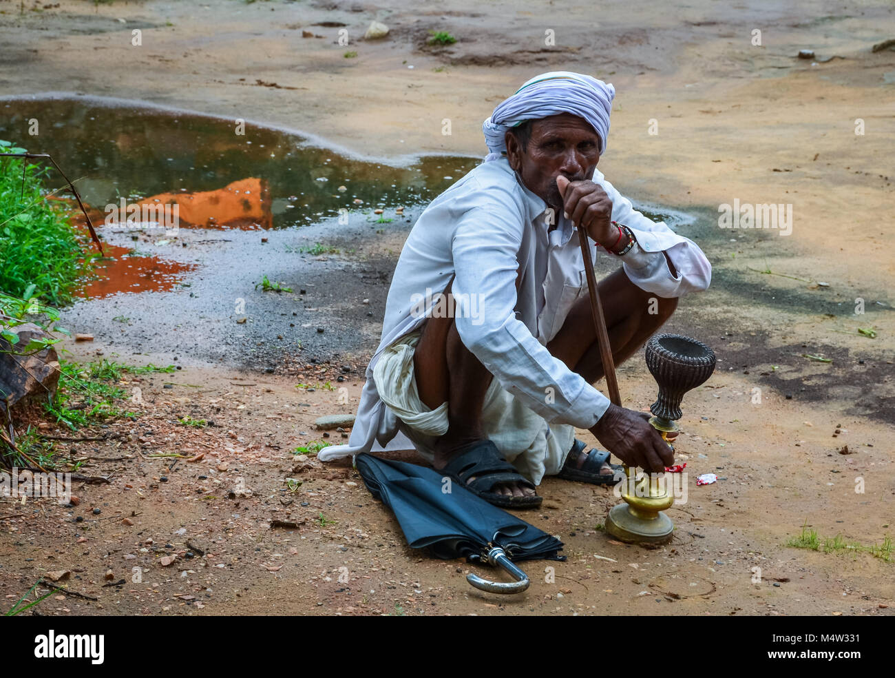 Indian male smoking a hookah while squatting on the ground in a rural ...