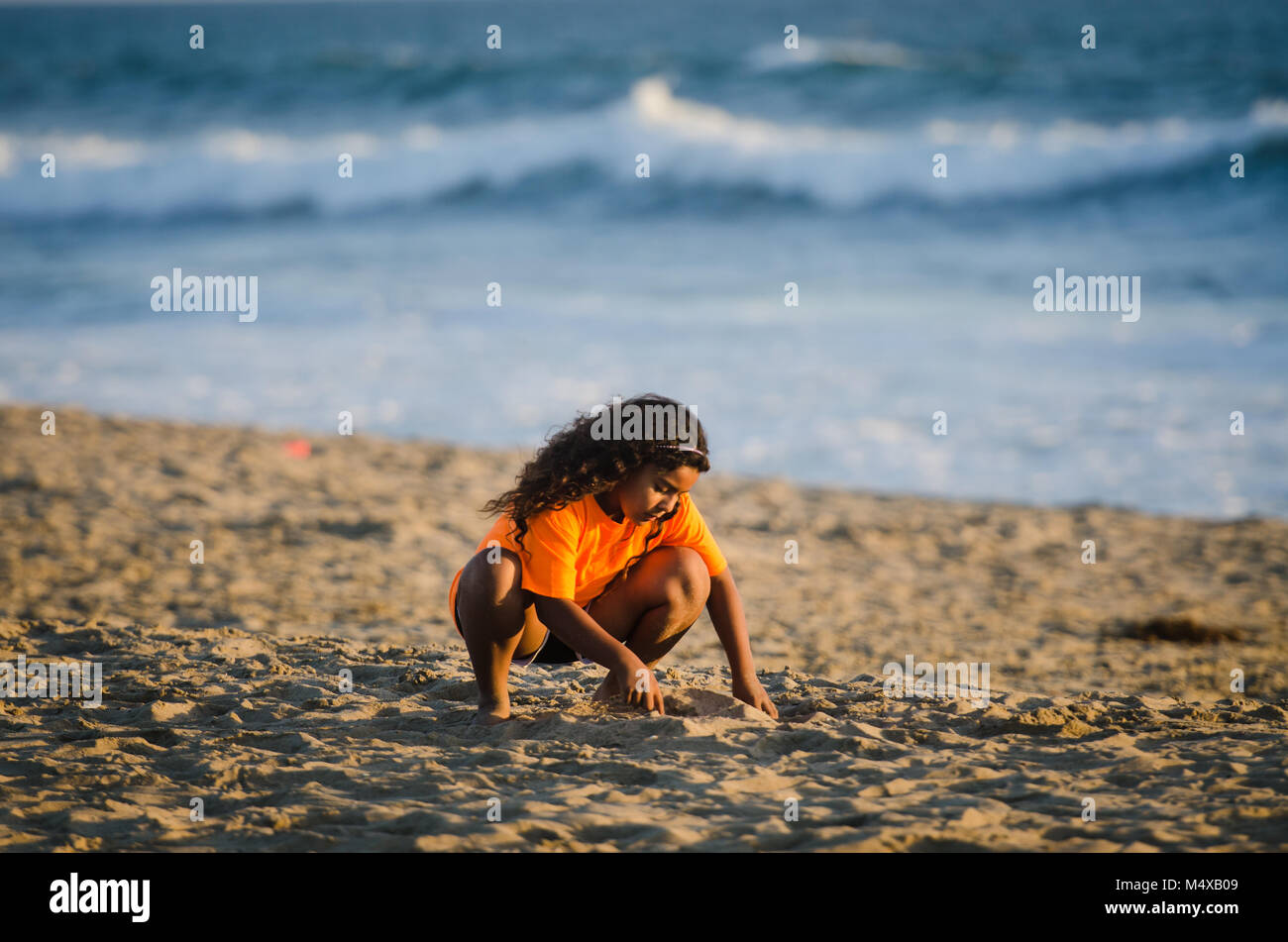 Multiracial girl with long curly hair digging in the sand on the shore ...