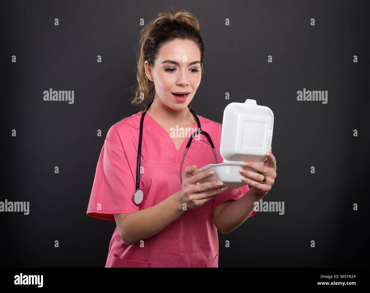 Portrait of beautiful young doctor opening lunch box looking surprised ...