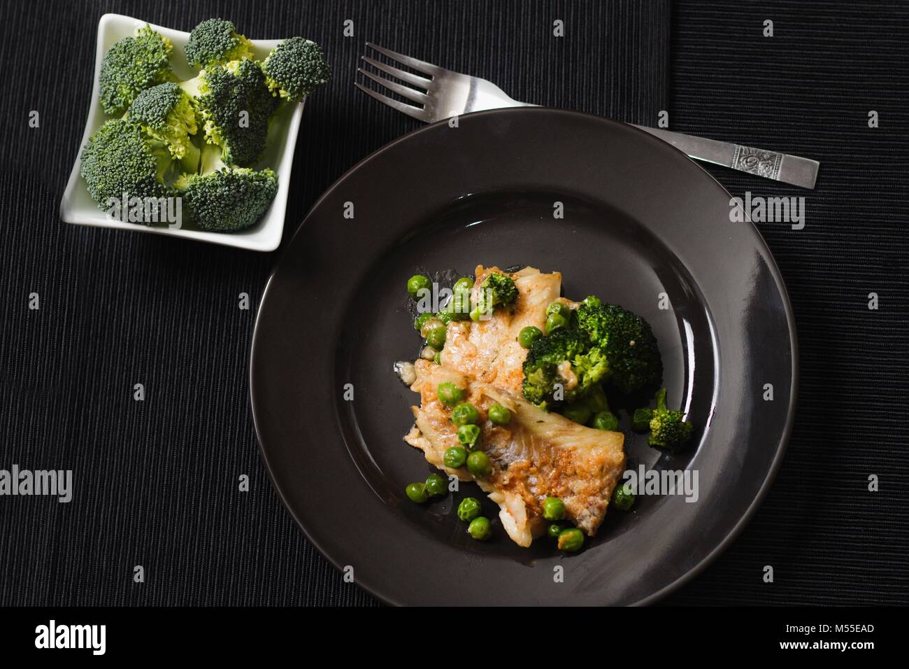 Top down view of serving of fried fish with peas and broccoli on black ...