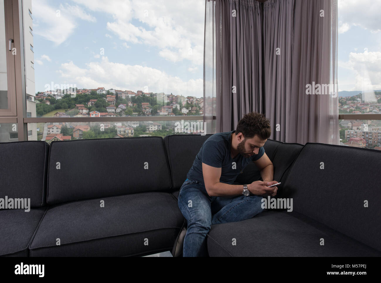 young man sitting on sofa and using a mobile phone near the window at ...