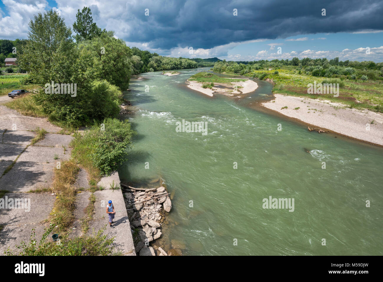 Man fishing in Prut river under dark clouds seen from highway bridge ...