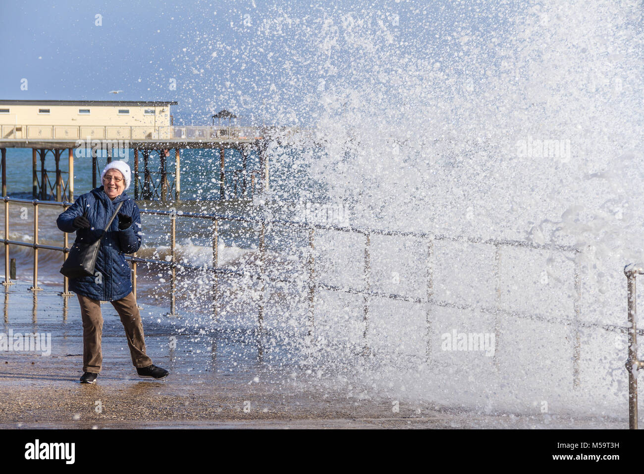 A woman avoiding being splashed by a wave at high tide on the promenade ...
