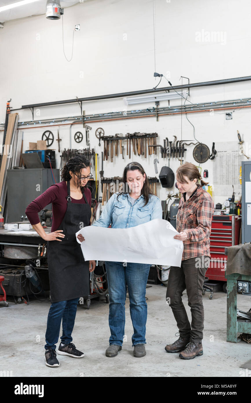 Three women standing in metal workshop, holding technical blueprint ...