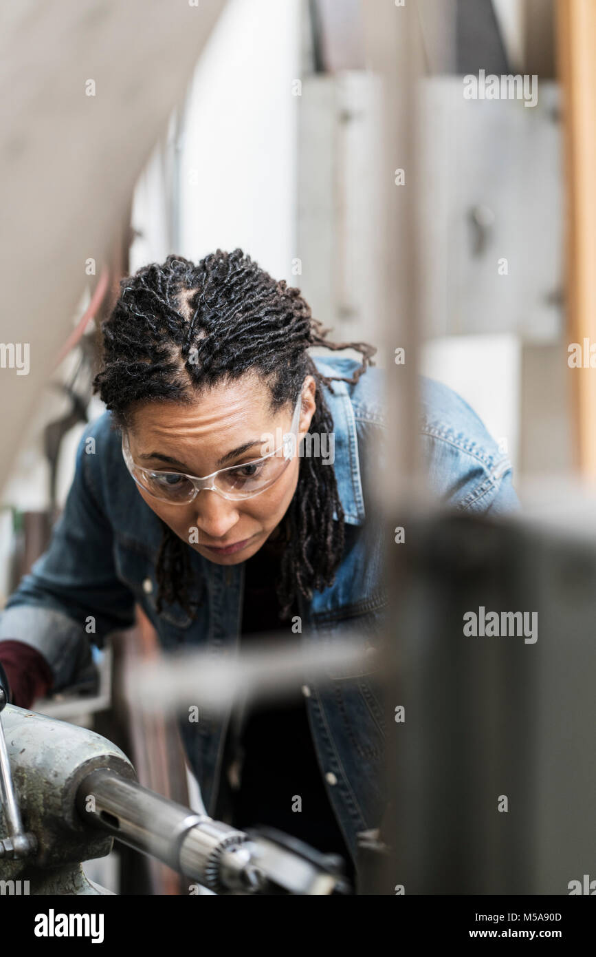 Woman wearing safety glasses standing in a metal workshop, working at a ...