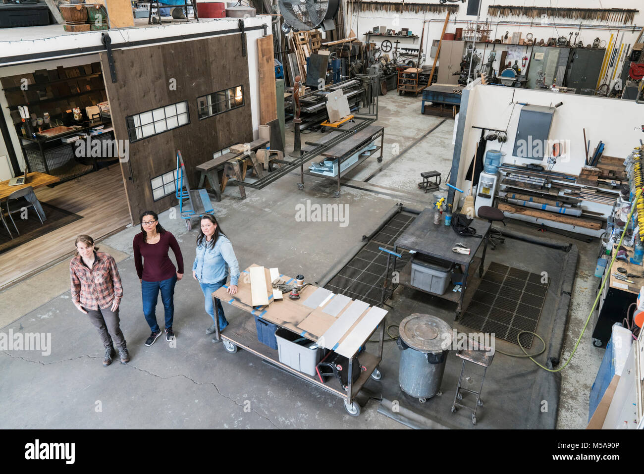 High angle view of three women standing in metal workshop, looking at ...
