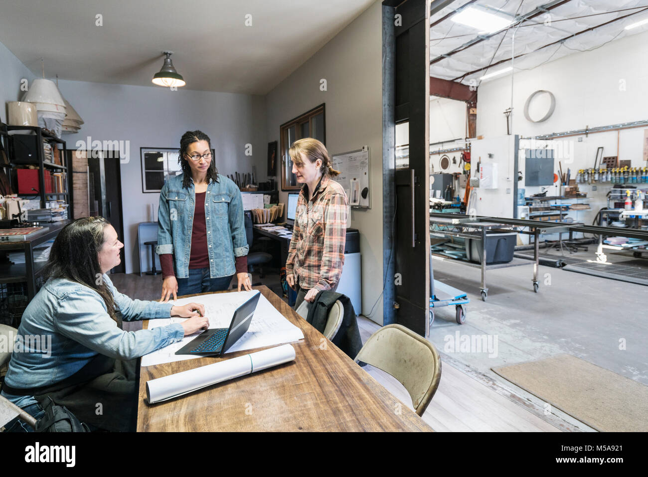Three women gathered around table in office area of a metal workshop ...