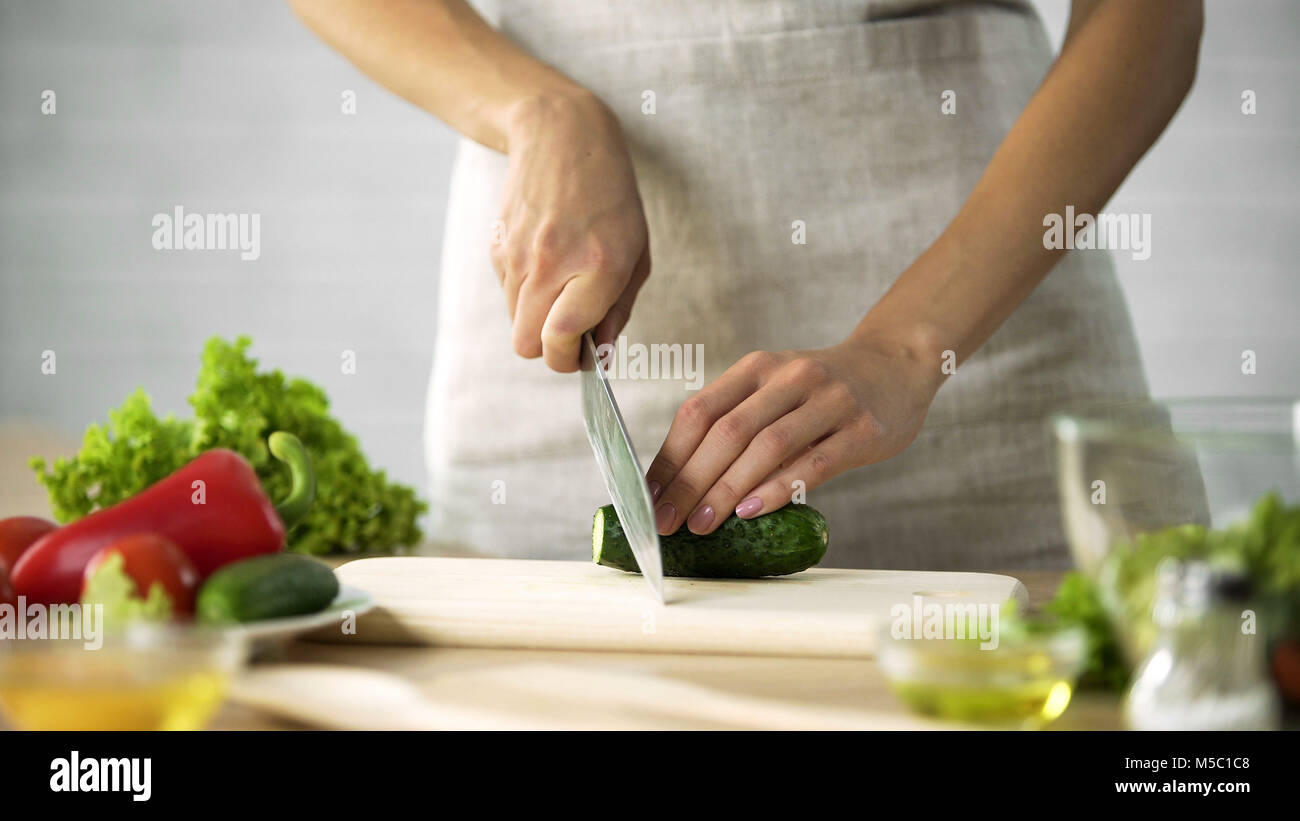 Female chef slicing fresh cucumber with a knife on wooden board ...