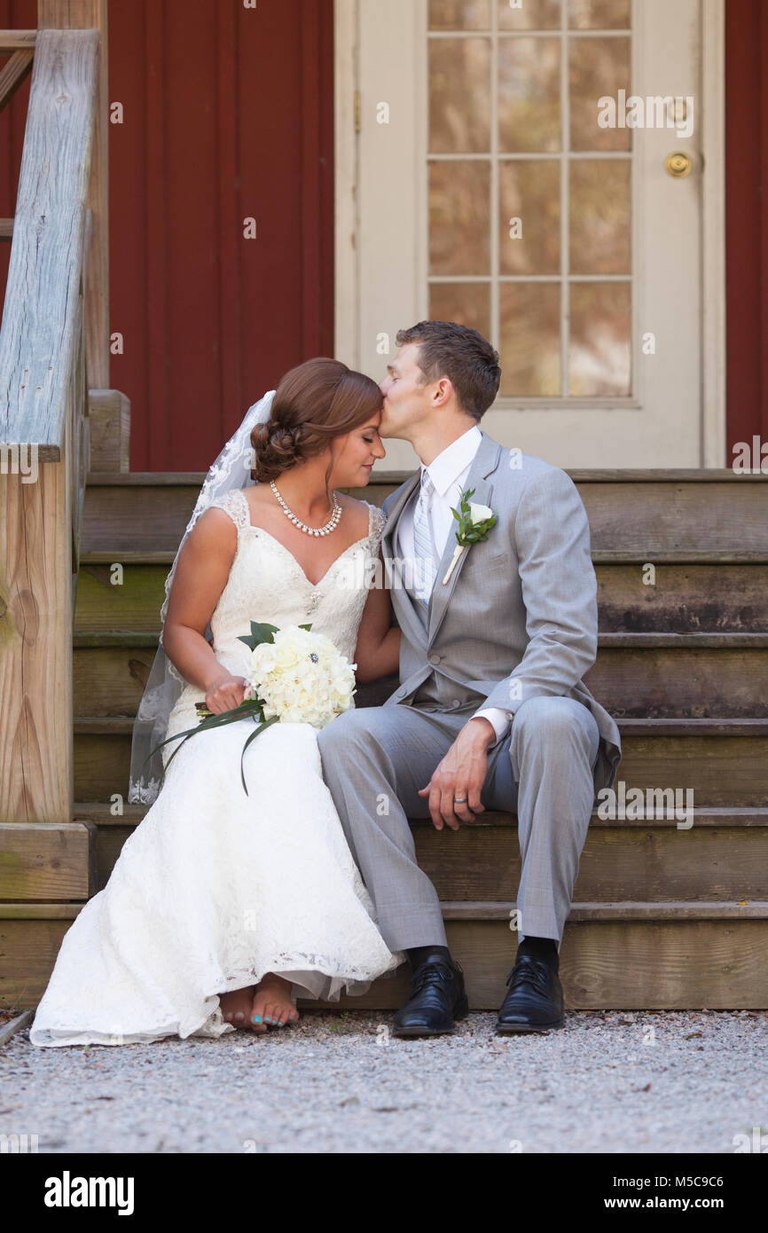 Young newly married couple seated on steps, groom kissing bride on ...