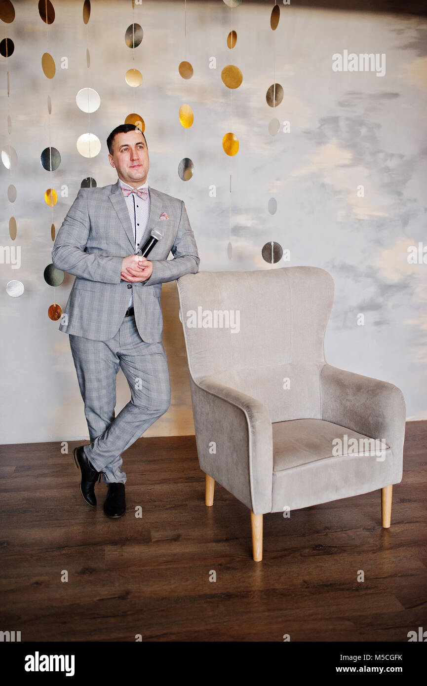 Handsome man in gray suit with microphone against chair background on ...