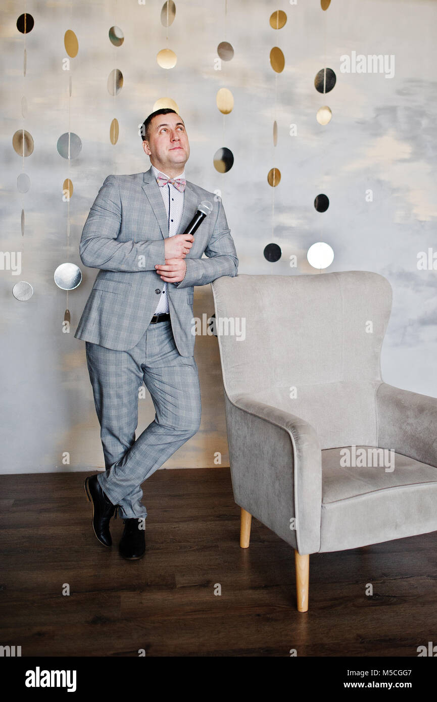 Handsome man in gray suit with microphone against chair background on ...