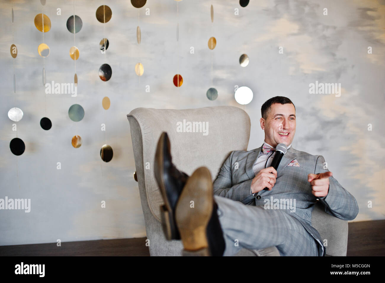 Handsome man in gray suit with microphone sitting on chair background ...