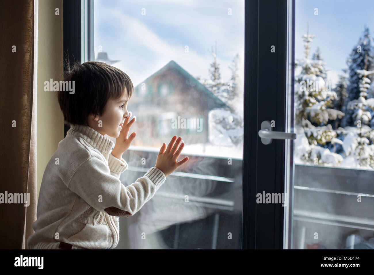 Preschool boy, sitting by the window in living room, looking at a snowy ...