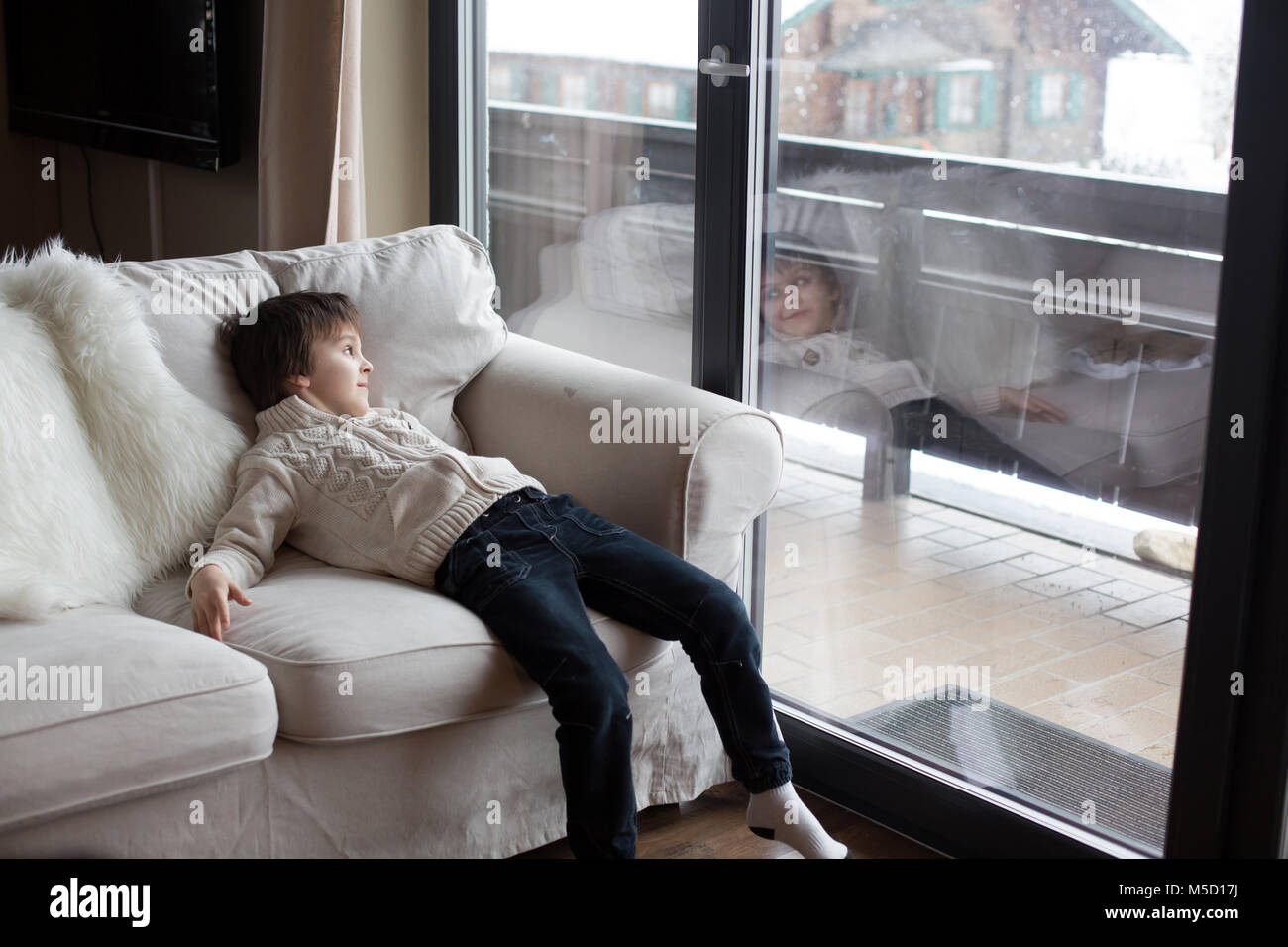 Preschool boy, sitting by the window in living room, looking at a snowy ...