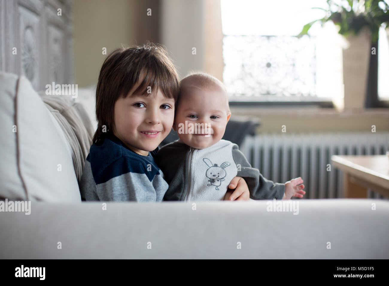Little baby boy and his older brother, sitting on a couch in sunny ...