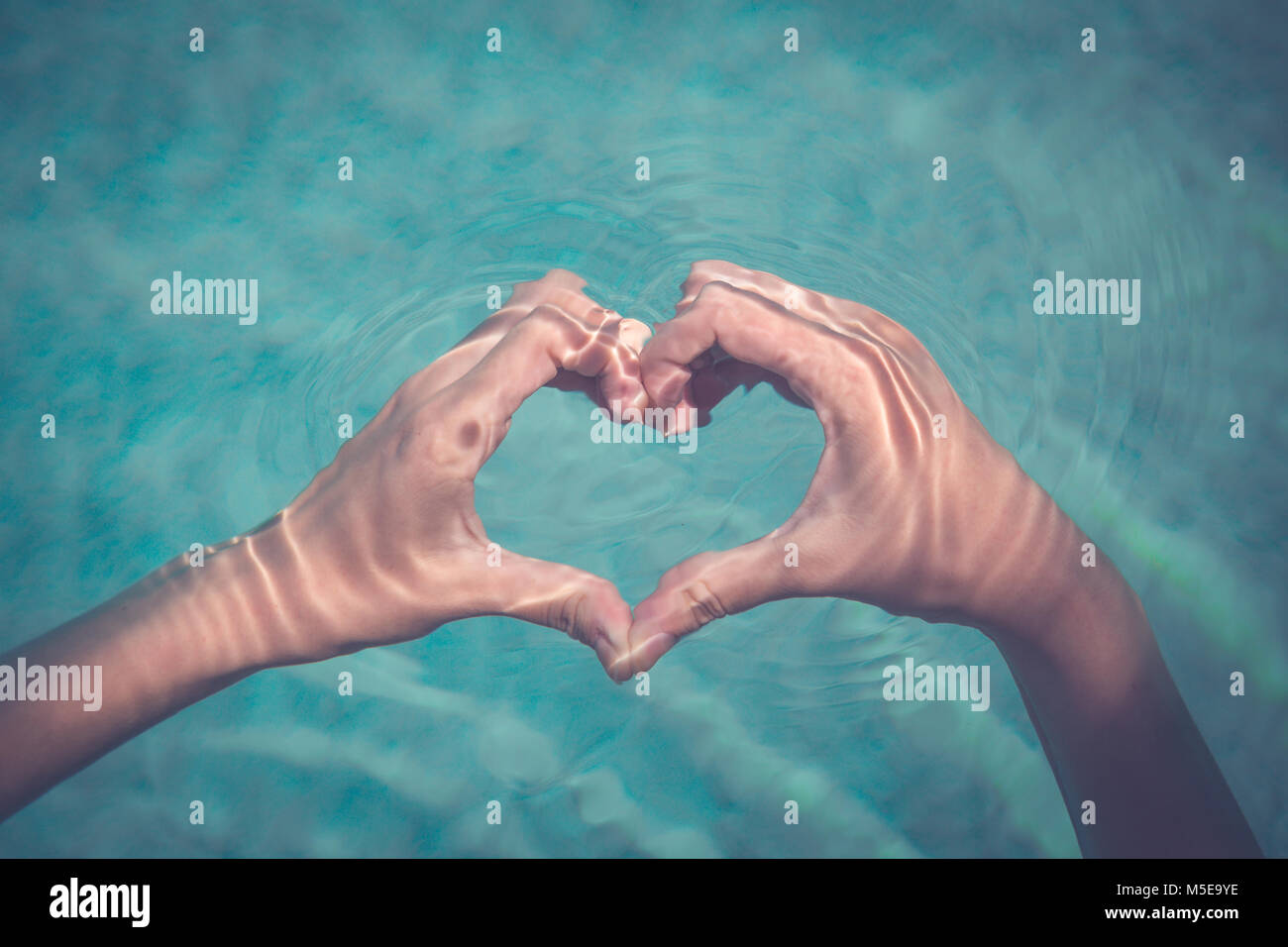 Close up of female hands creating heart shape on blue water surface ...