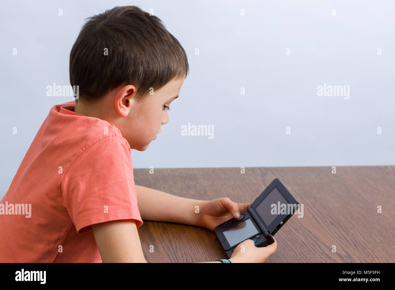 Young boy playing handheld video game at a table. Against grey ...